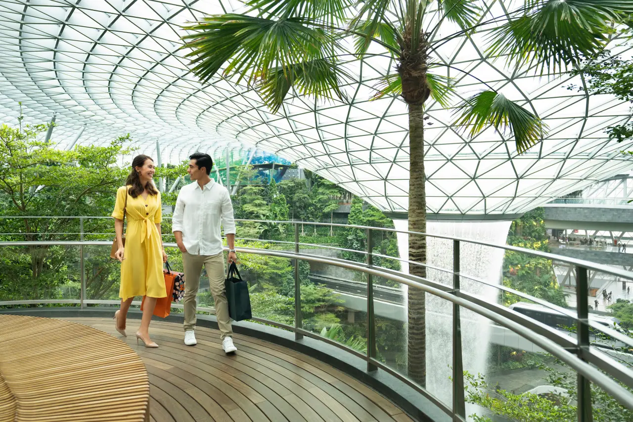 A couple walks along an indoor garden walkway at Jewel Changi Airport in Singapore, with lush greenery and the Rain Vortex waterfall in the background.