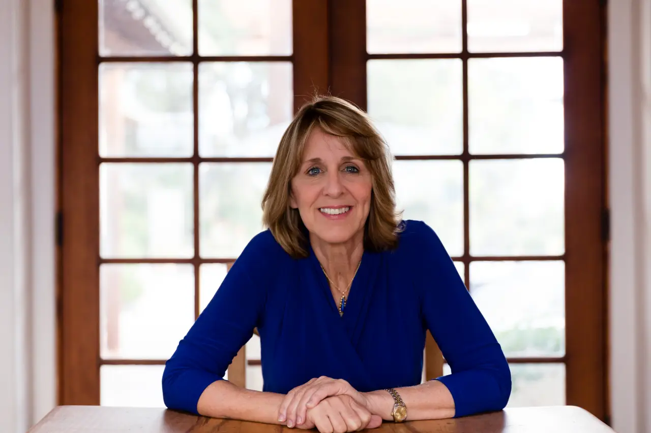 Diane Petras, retiring president of The Travel Institute, poses at a wooden table in front of windowed doors.